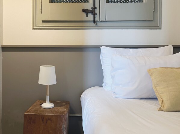 Cama bem arrumada com almofadas brancas e bege, mesa de cabeceira de madeira com abajur branco, e armário fixado na parede acima da cama.