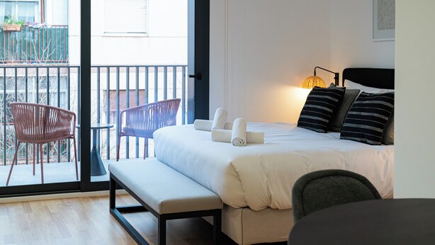 Um quarto com uma cama, um banco, duas cadeiras, uma mesinha de cabeceira e um abajur.