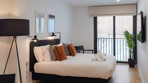 Quarto moderno com cama preta, roupa de cama branca e uma janela grande com vista para um edifício de tijolos.