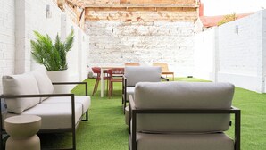 Uma área de estar moderna ao ar livre, com parede de tijolos brancos, grama sintética verde e um muro de pedra ao fundo.