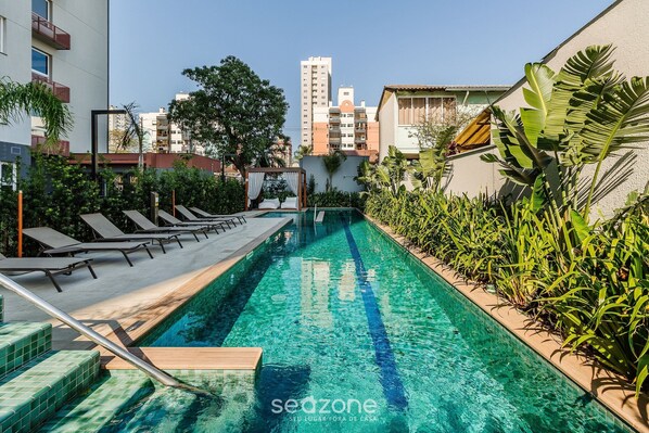 Piscina com espreguiçadeiras, cercada por vegetação e edifícios.