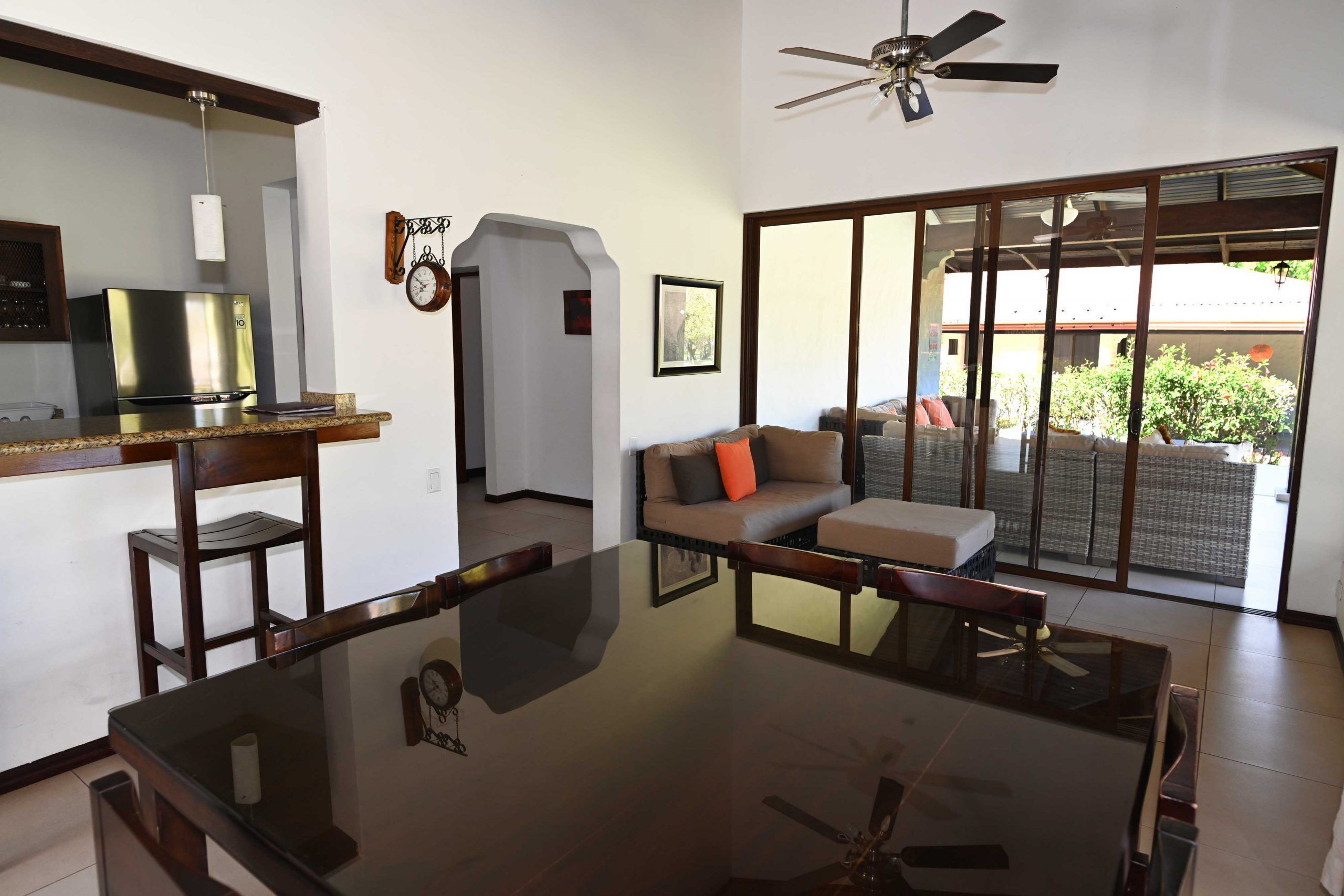 Uma sala de estar moderna com uma mesa de jantar de vidro, um sofá com almofadas, uma cozinha com geladeira e um ventilador de teto.