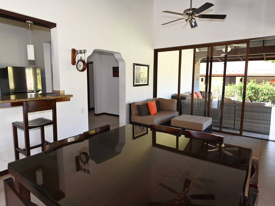 Uma sala de estar moderna com uma mesa de jantar de vidro, um sofá com almofadas, uma cozinha com geladeira e um ventilador de teto.