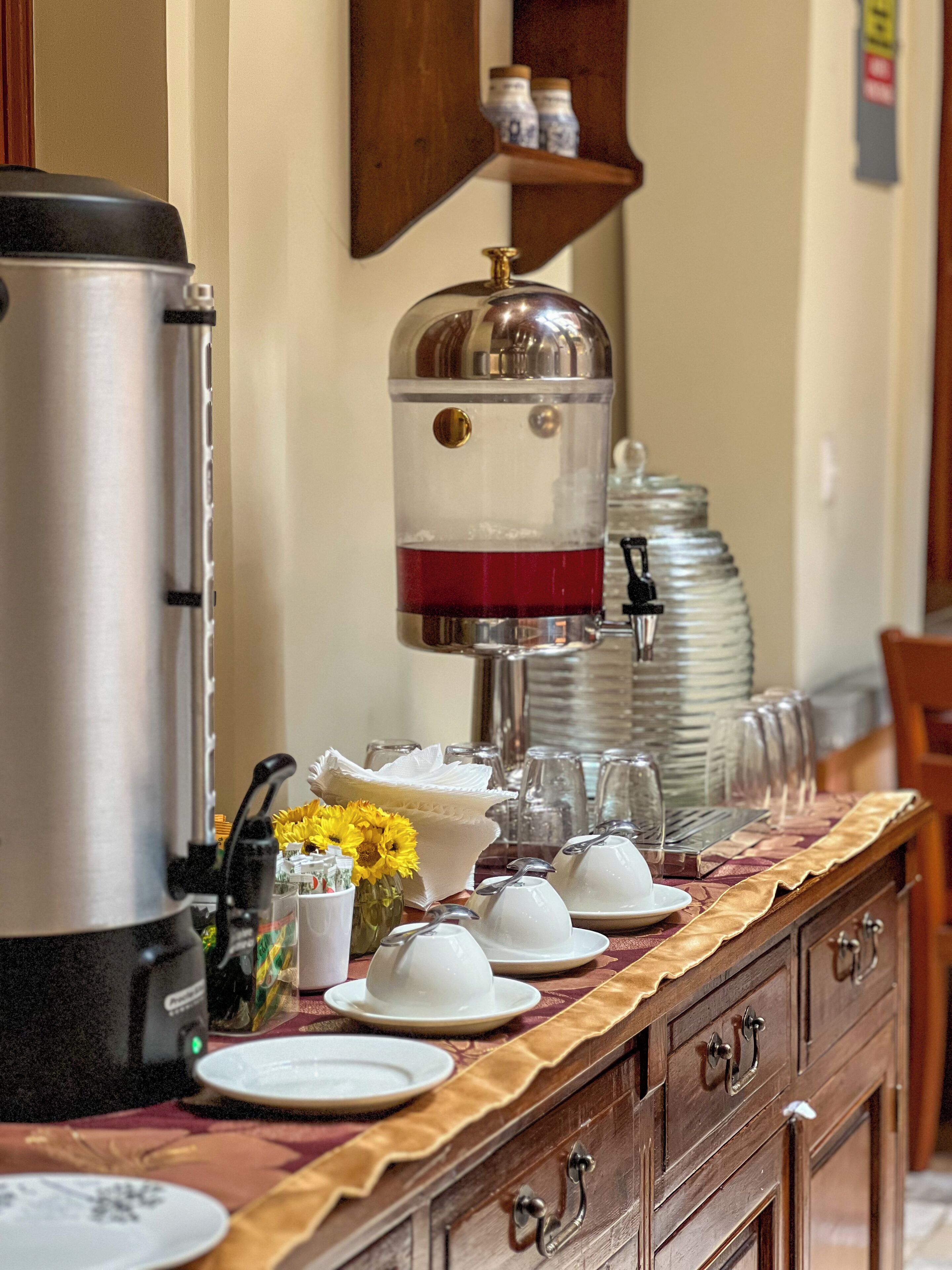 Disposi ção de buffet com máquina de café, dispensador de água e copos empilhados sobre uma mesa de madeira.