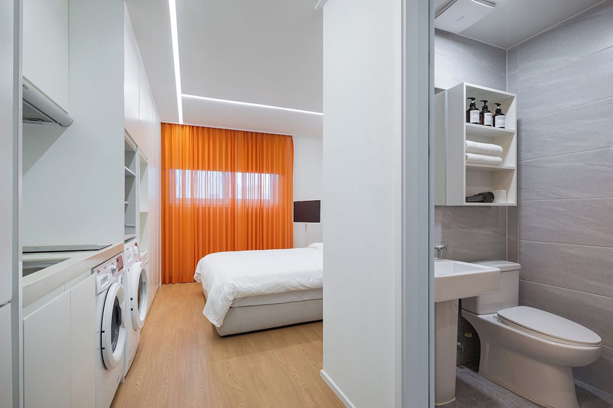 Quarto de hotel compacto com cama, máquina de lavar, pia e vaso sanitário.