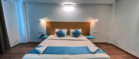 Cama bem arrumada com detalhes azuis, duas mesas de cabeceira com abajures e cabeceira com iluminação embutida.