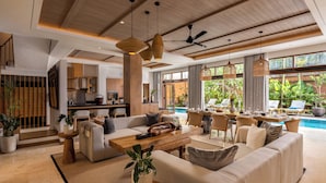 Ampla sala de estar com um sofá grande, uma mesa de centro de madeira e uma mesa de jantar. É visível uma cozinha ao fundo, e uma piscina através das janelas.