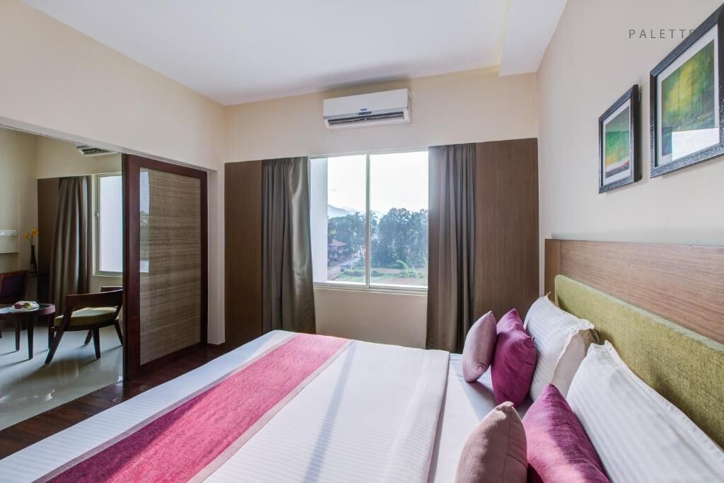 Quarto de hotel com cama, escrivaninha e cadeira. Há uma janela com cortinas, um aparelho de ar condicionado e quadros nas paredes.