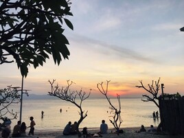 Cena de praia com pessoas sentadas e caminhando pela margem, árvores e um pôr do sol sobre a água.