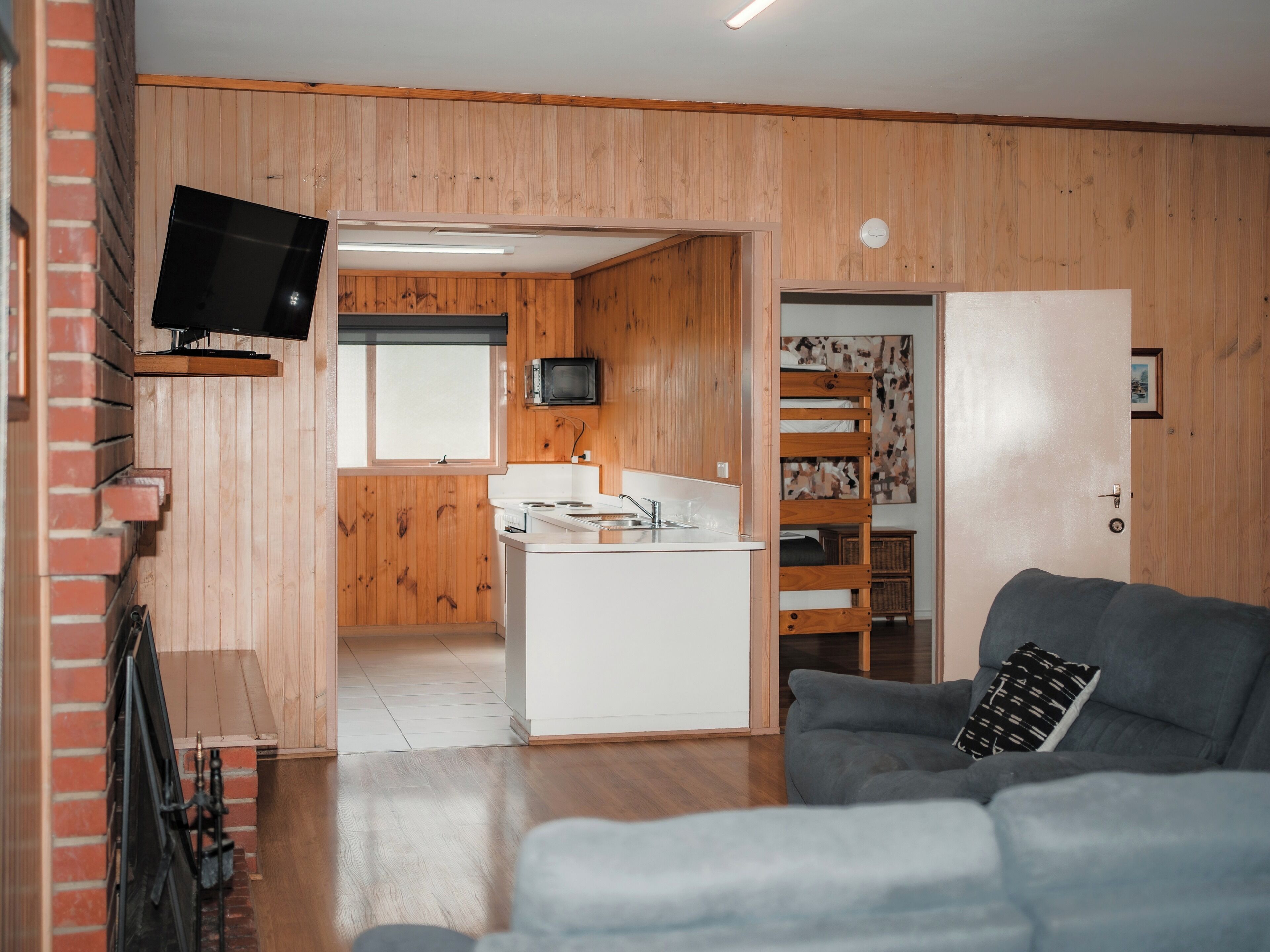 Uma sala de estar moderna com um sofá, uma televisão fixada em uma parede de tijolos, uma área de cozinha com micro-ondas e uma porta de madeira.