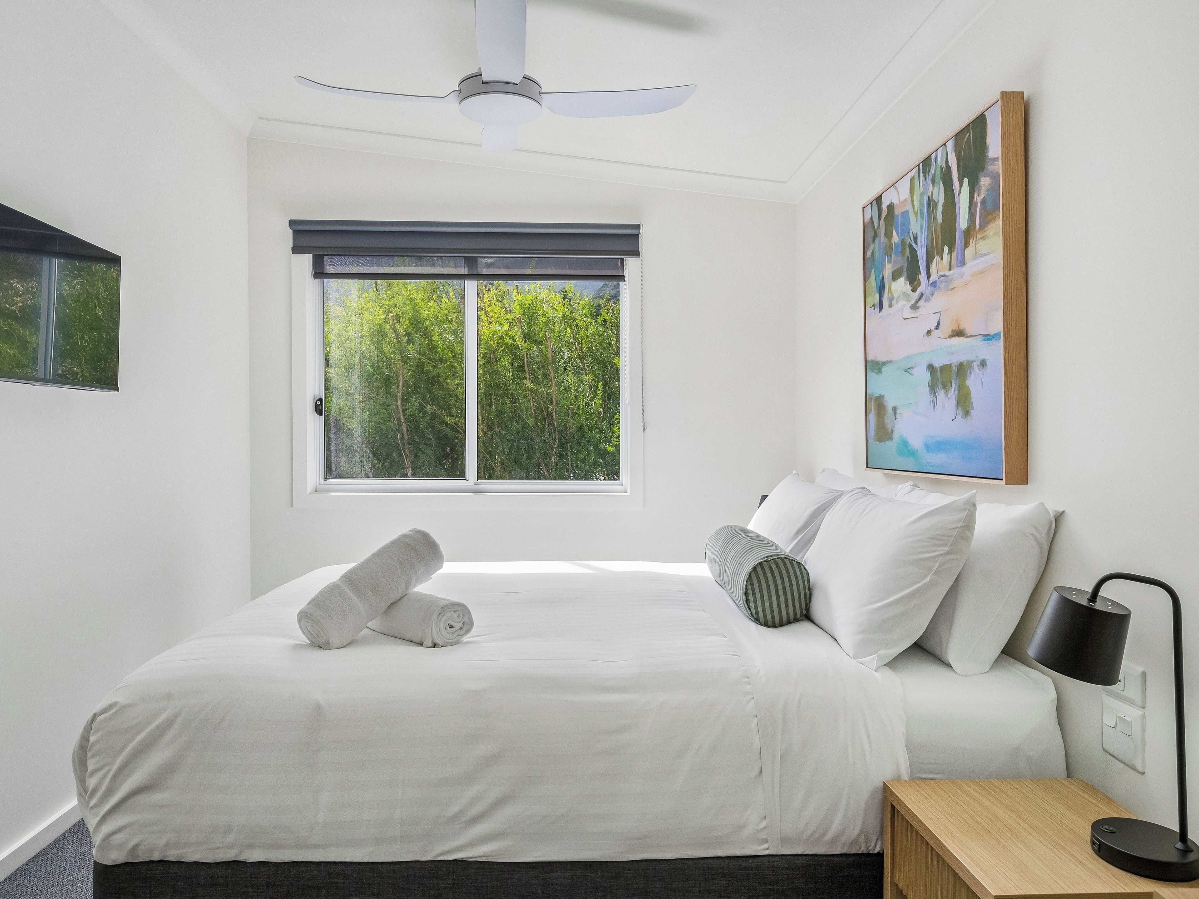 Quarto de hotel com cama, janela com vista para árvores, pintura na parede e mesa de cabeceira com abajur.