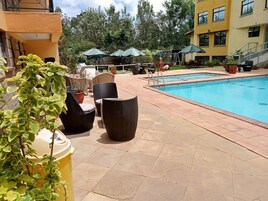 Uma área ao lado da piscina com cadeiras de vime e guarda-sóis, piscina e um edifício ao fundo.
