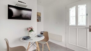 Uma sala de jantar com uma mesa branca, duas cadeiras, um vaso com flores e uma televisão de tela plana fixada na parede.