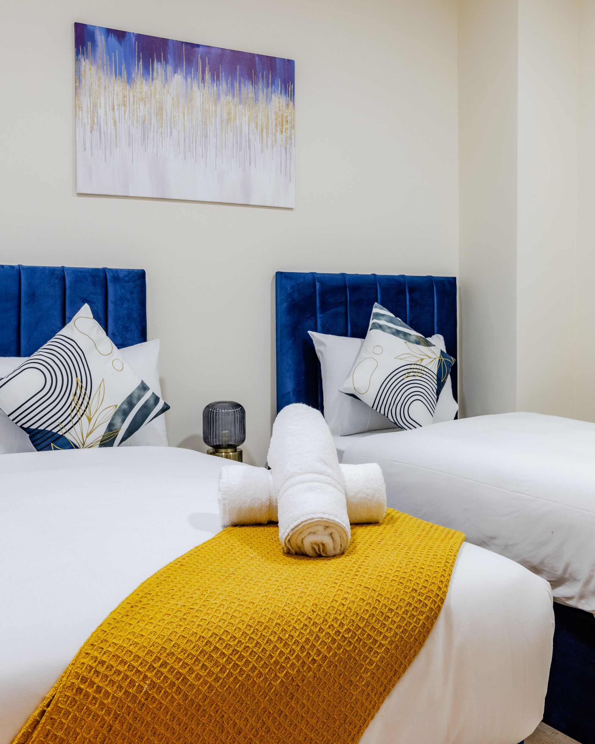 Quarto de hotel com duas camas, cada uma com cabeceira azul, roupa de cama branca e almofadas decorativas. Um cobertor amarelo está jogado sobre a cama em primeiro plano.