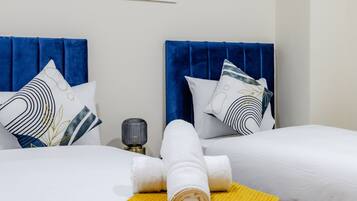 Quarto de hotel com duas camas, cada uma com cabeceira azul, roupa de cama branca e almofadas decorativas. Um cobertor amarelo está jogado sobre a cama em primeiro plano.