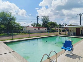 Piscina com cadeira azul, escada e área coberta com cadeiras e um aviso.