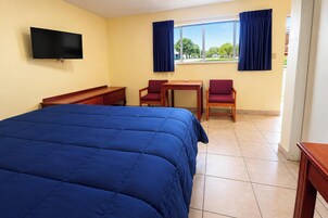 Quarto de hotel com uma cama grande, uma escrivaninha, duas cadeiras, uma televisão e uma janela com cortinas.