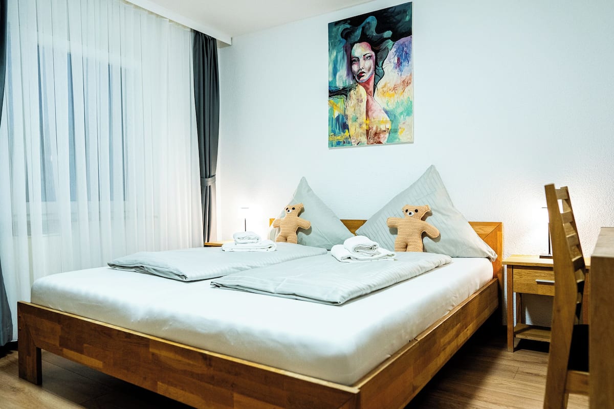 Cama bem arrumada com travesseiros e um ursinho de pelúcia, uma escrivaninha de madeira e um quadro na parede.