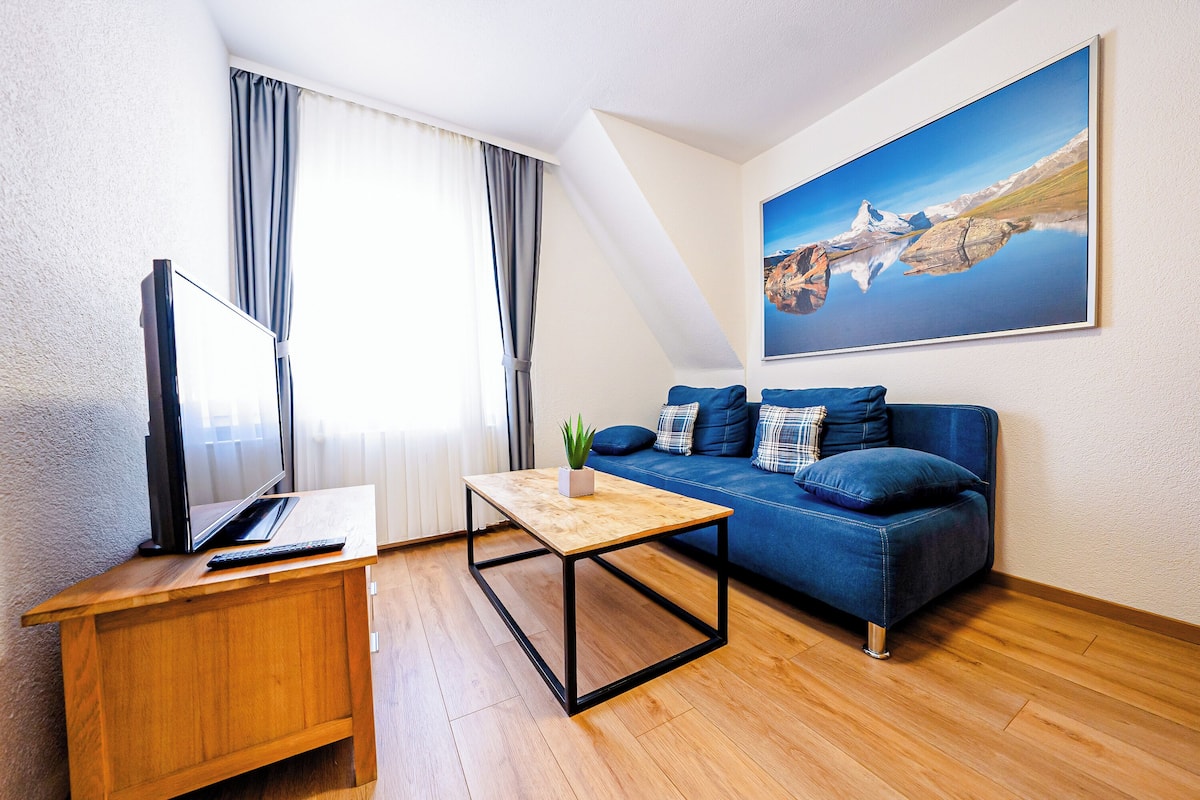 Uma sala de estar moderna com uma mesa de centro de madeira, um sofá azul com almofadas, uma TV de tela plana em um suporte de madeira e um grande quadro com paisagem na parede.