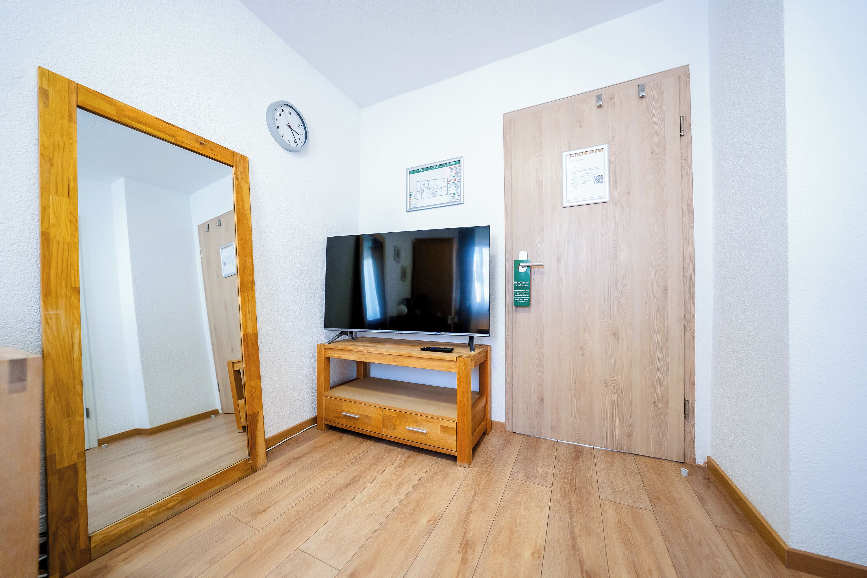 Quarto com um suporte de TV de madeira, um relógio na parede e uma porta com uma placa.
