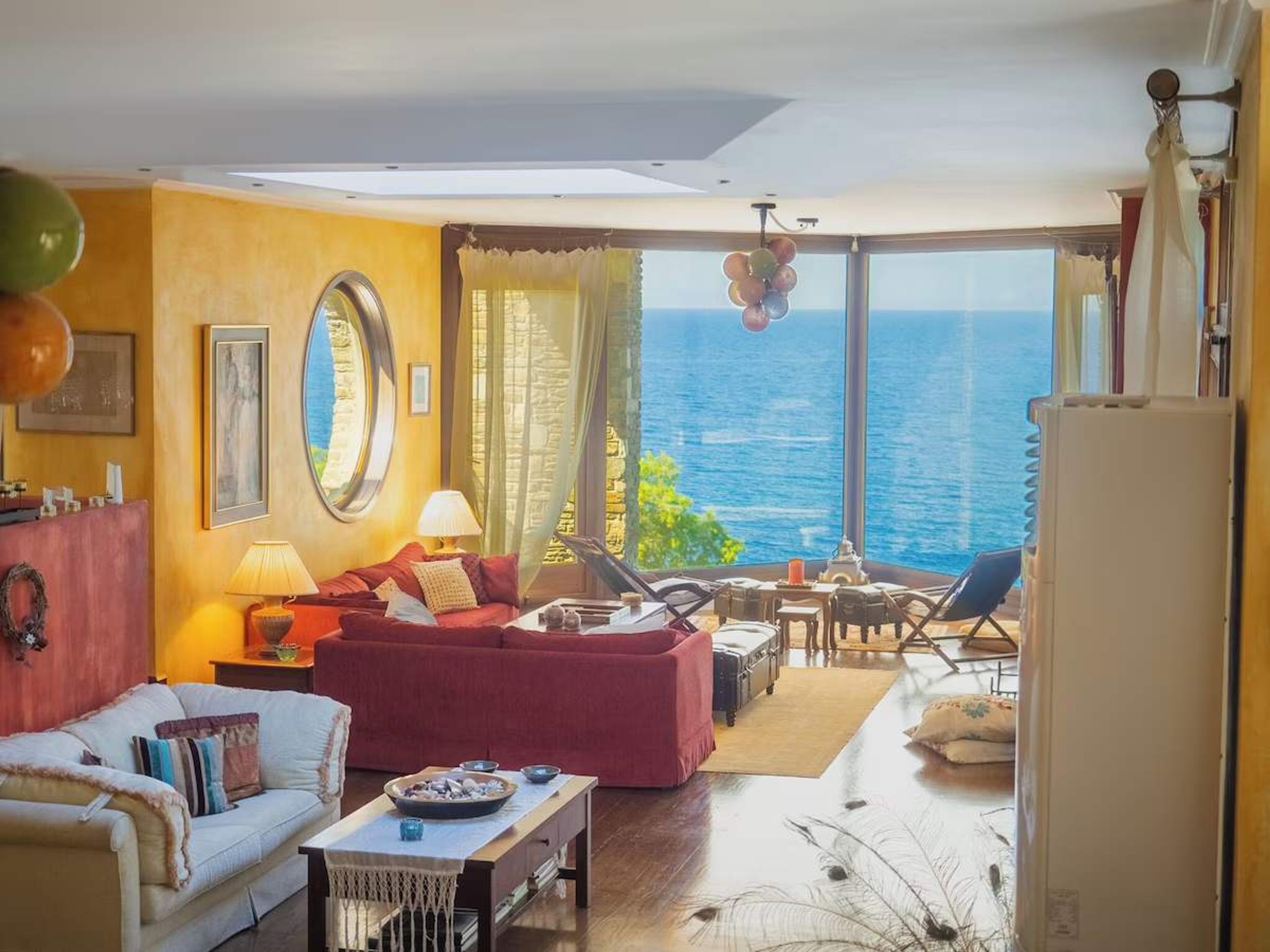 Uma sala de estar espaçosa com uma grande janela com vista para o mar, um sofá vermelho, um sofá branco e uma mesa de jantar.