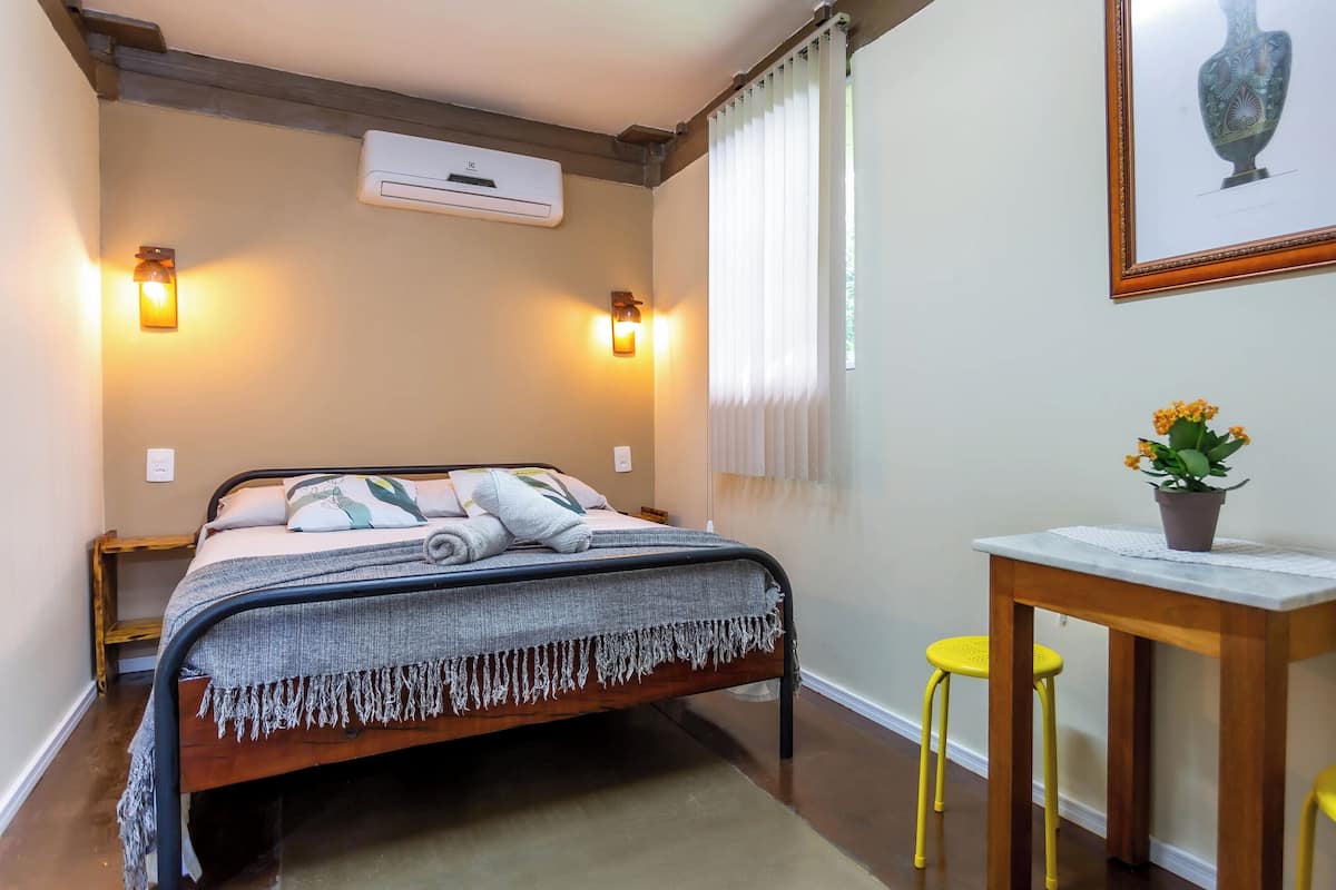 Cama bem arrumada com manta, duas luminárias de cabeceira, uma mesinha com uma planta e um banquinho amarelo.