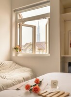 Quarto de hotel com uma cama, uma janela com vista para um edifício alto e uma mesa de cabeceira com um vaso de flores.