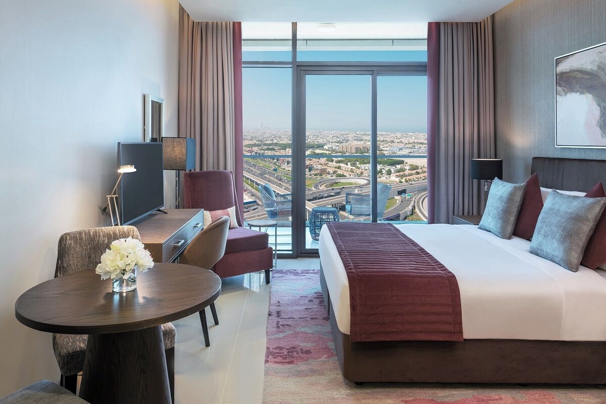 Quarto de hotel moderno com uma cama grande, uma escrivaninha e vista para a paisagem urbana por janelas do chão ao teto.