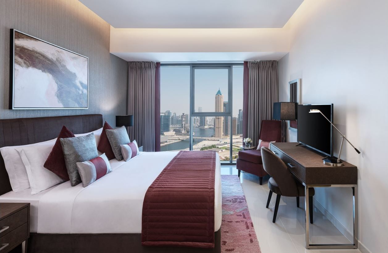 Quarto de hotel moderno com uma cama grande, escrivaninha, televisão e vista para a cidade através de amplas janelas.