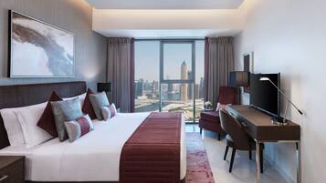 Quarto de hotel moderno com uma cama grande, escrivaninha, televisão e vista para a cidade através de amplas janelas.