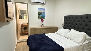 Quarto com cama, mesa de cabeceira, espelho, ar condicionado fixado na parede e uma pintura de paisagem.