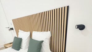 Cama bem arrumada com lençóis brancos e duas almofadas verdes, cabeceira de ripas de madeira, mesa de cabeceira pequena de madeira e interruptor de luz fixado na parede.