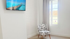 Quarto com piso de madeira, parede branca, janela com cortinas e uma cadeira com padrão floral.