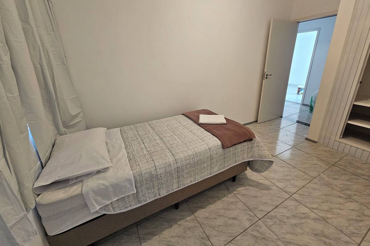 Uma cama pequena e bem arrumada, com um cobreleja estampado e uma toalha branca dobrada.