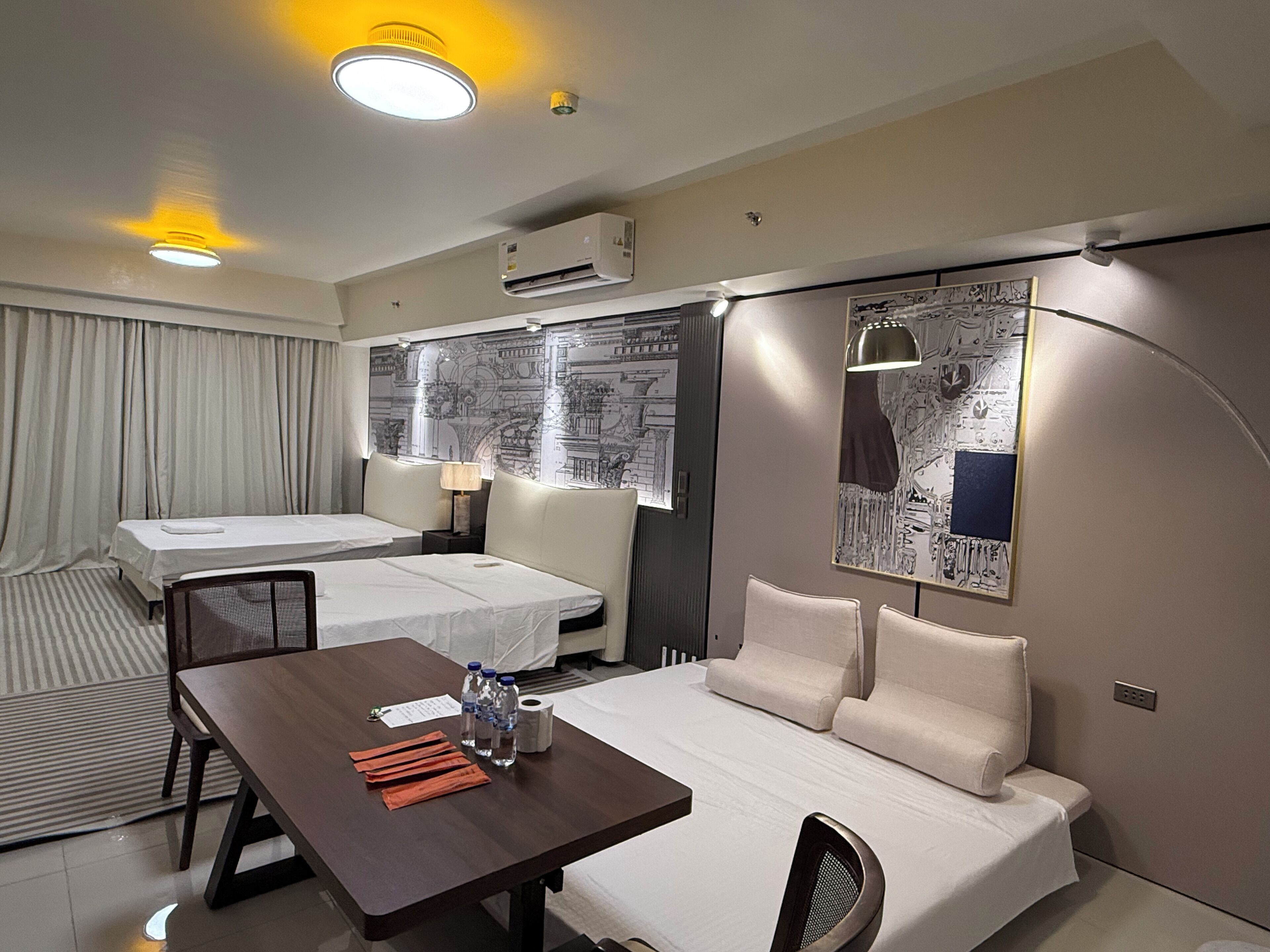 Quarto de hotel com cama, escrivaninha, duas cadeiras, mesa e uma janela grande.
