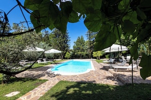 Piscina com ladrilhos de pedra, rodeada por grama e árvores, e com guarda-sóis para sombra.