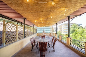 Um pátio externo com mesa e cadeiras de madeira, teto de palha e vista para prédios e árvores.