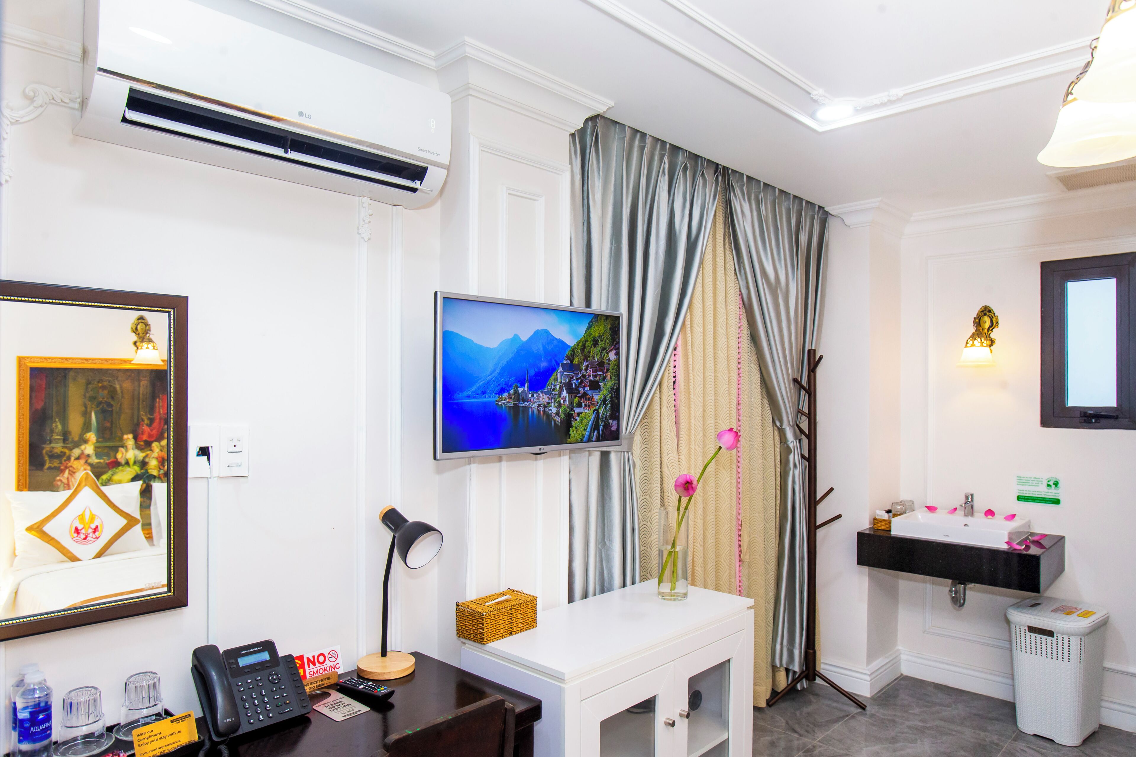 Quarto de hotel com televisão de tela plana, uma escrivaninha com telefone, luminária, cesta, vaso com flores, pia e espelho.