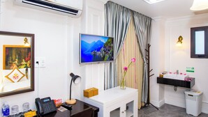 Quarto de hotel com televisão de tela plana, uma escrivaninha com telefone, luminária, cesta, vaso com flores, pia e espelho.
