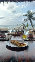 Uma mesa com um prato de comida, um copo de água e um copo de suco, com vista para a praia, palmeiras e o oceano ao fundo.