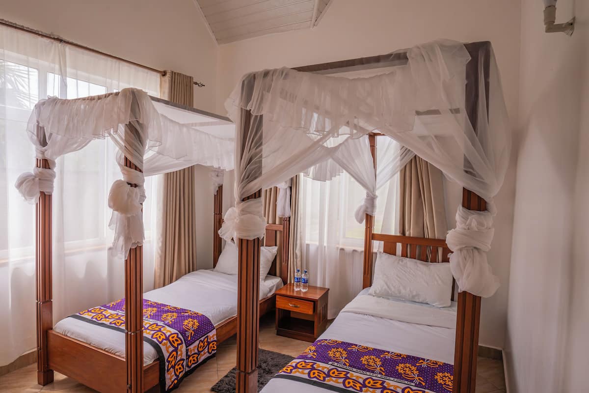 Quarto com duas camas de madeira, cada uma com dossel e colcha colorida. Há uma mesinha de cabeceira entre as camas com uma garrafa de água.