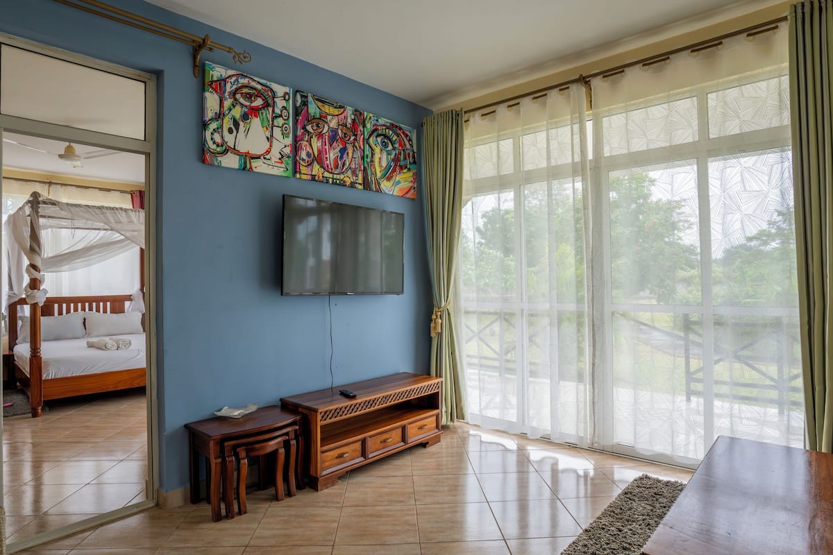Quarto com cama, televisão, mesa consola de madeira e uma janela grande com cortinas.