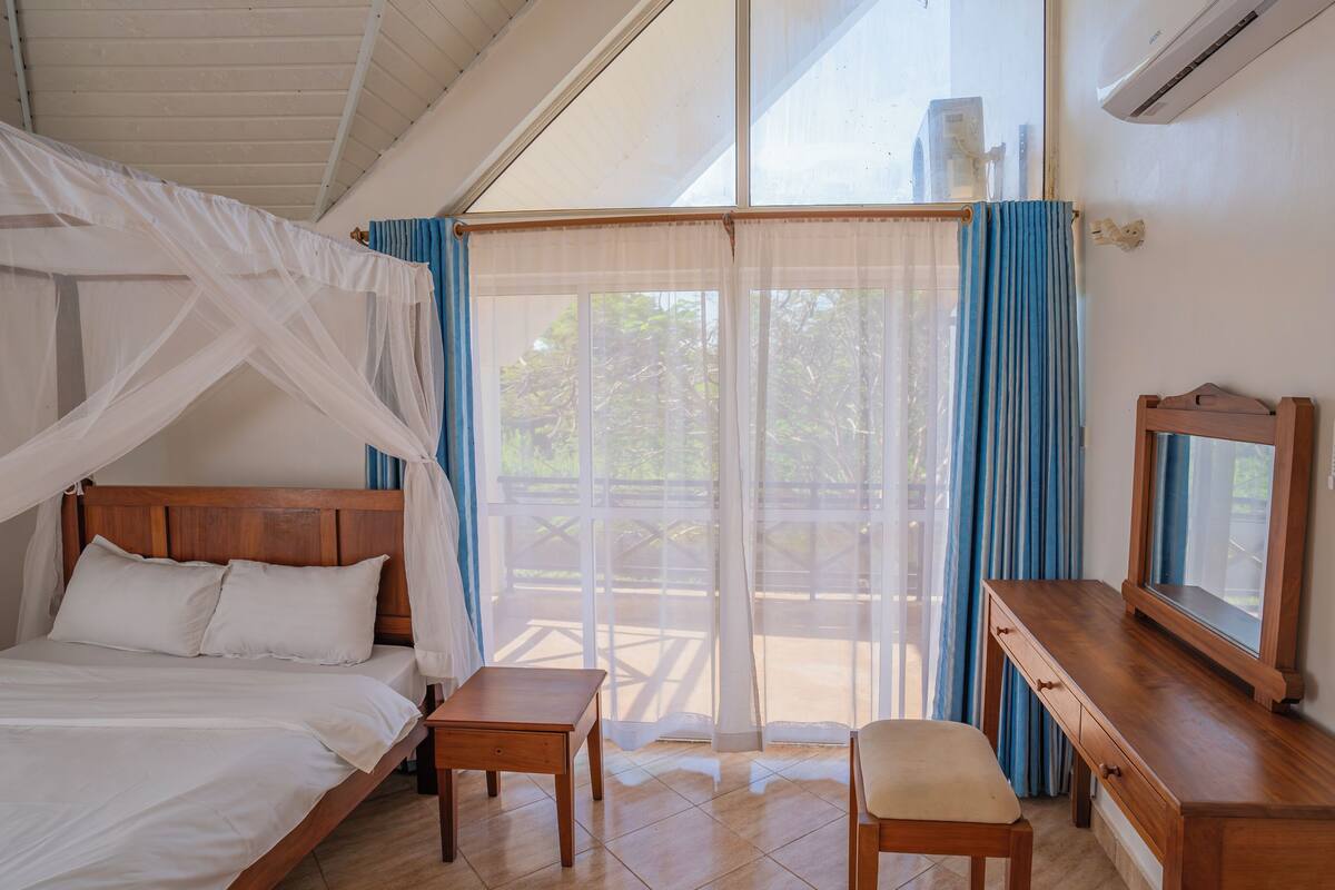 Quarto com cama, mesa de cabeceira, cadeira e escrivaninha com espelho. Há uma janela com cortinas e um ar condicionado na parede.