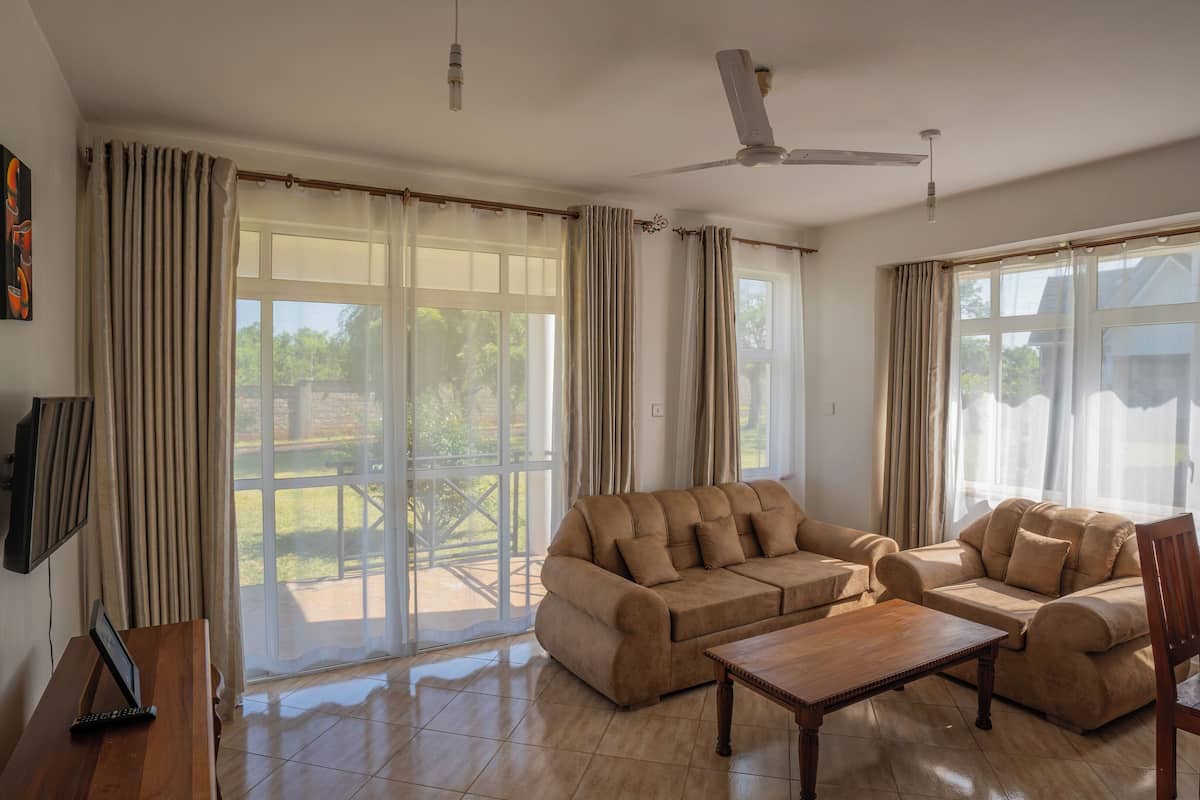 Uma sala de estar espaçosa com sofá seccional, mesa de centro de madeira e televisão de tela plana. Grandes janelas com cortinas oferecem bastante luz natural e vista para o exterior.