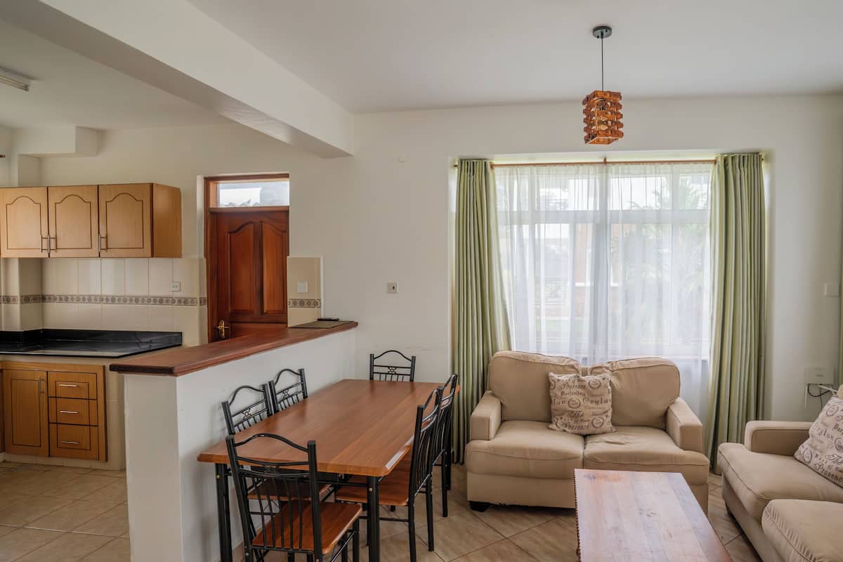 Uma sala de estar com uma mesa de jantar de madeira, sofá bege e uma mesa de centro. Há uma área de cozinha com armários de madeira e bancada preta.