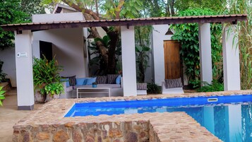 Uma área de piscina com paredes de pedra, uma pérgula de madeira, um sofá com almofadas e um sinal indicando banheiro e chuveiro.