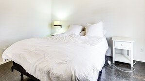 Cama bem arrumada com edredom branco e travesseiros, mesa de cabeceira com abajur e tomada na parede.