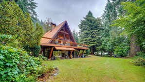 Uma cabana com telhado inclinado, deck de madeira e uma grande janela com vista para um gramado.