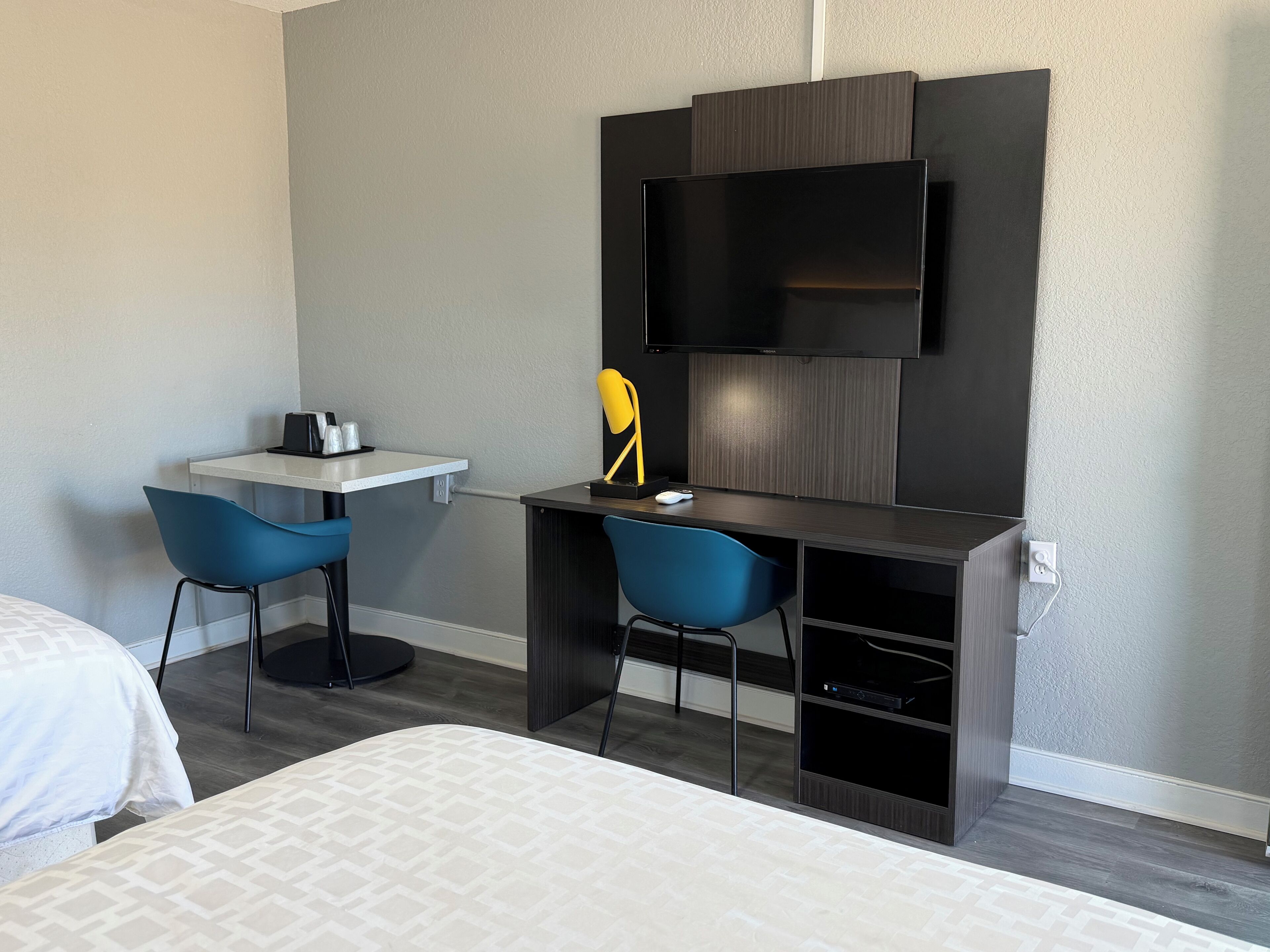 Quarto de hotel com uma cama, uma escrivaninha com duas cadeiras, uma televisão de tela plana na parede e uma pequena mesa com cafeteira e saleiro e pimenteiro.