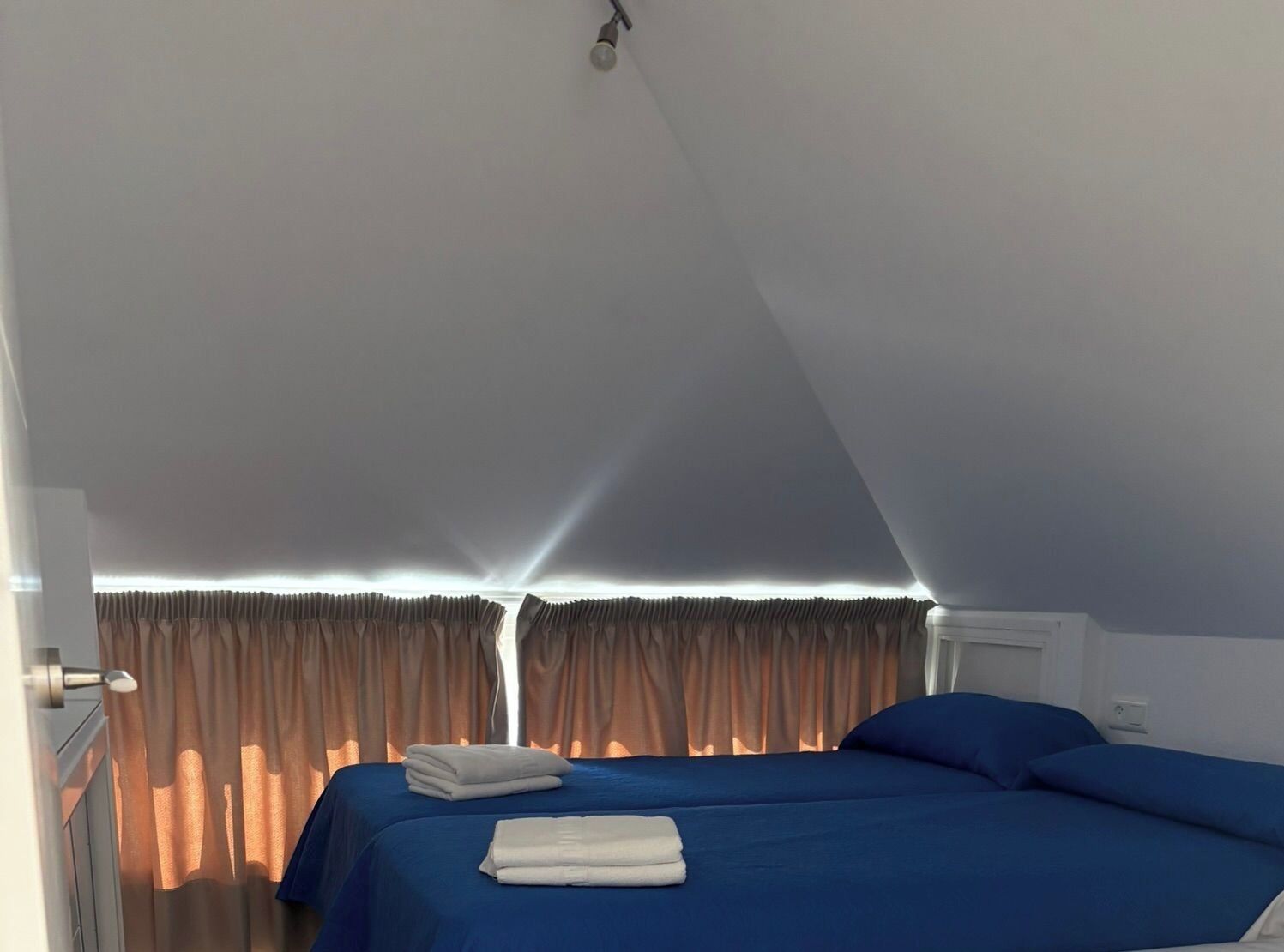 Quarto com teto inclinado, cama com roupa de cama azul e cortinas.